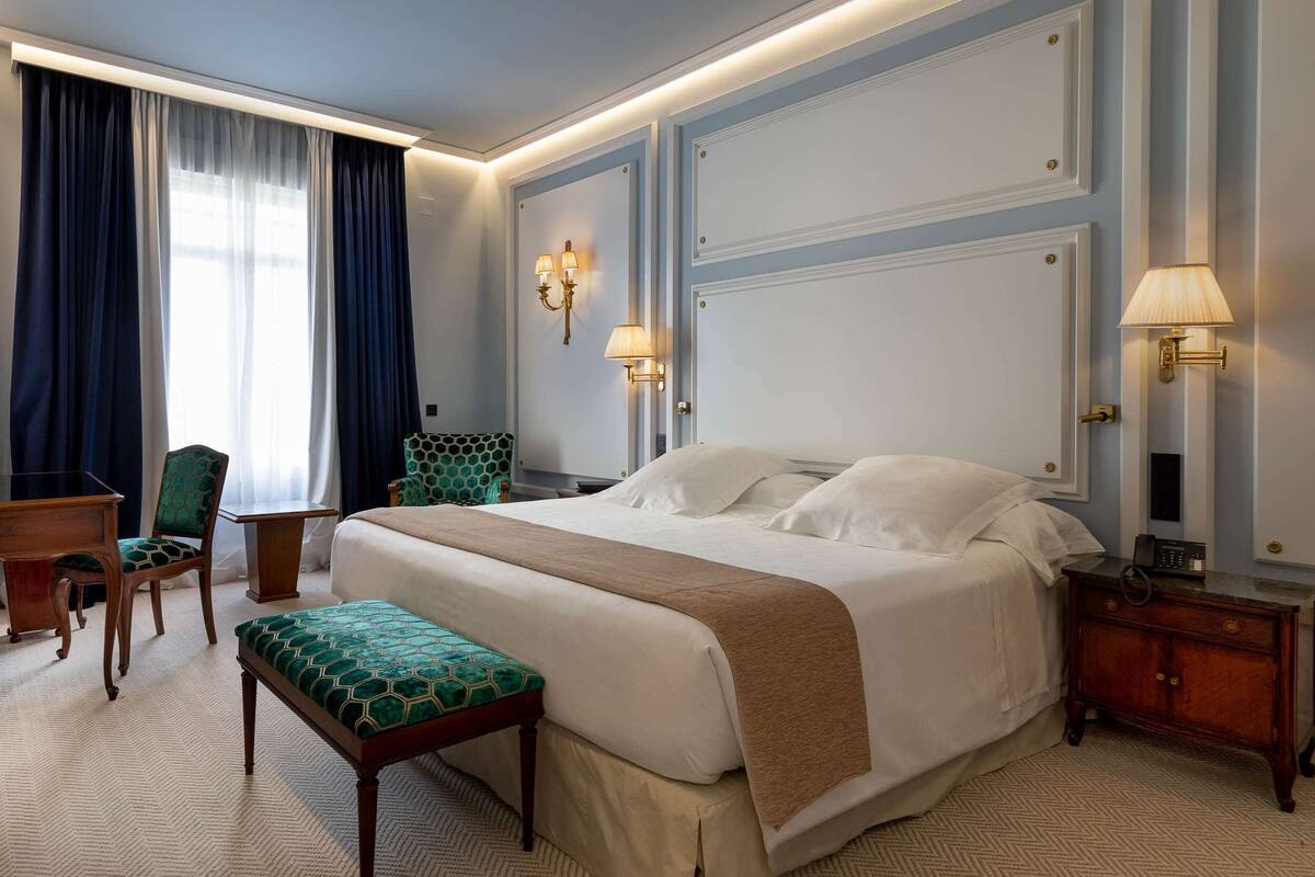 Quarto de hotel com uma cama grande, uma escrivaninha, uma cadeira e duas arandelas.