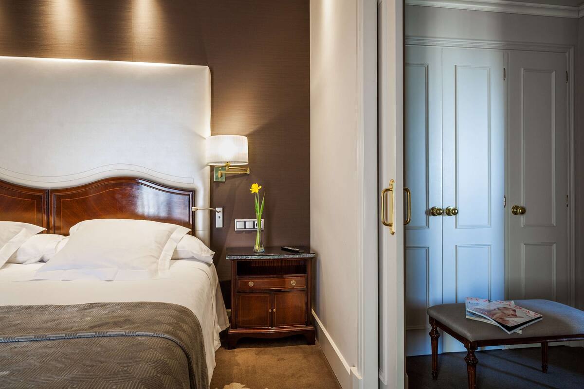 Quarto de hotel com uma cama grande, uma mesa de cabeceira com abajur, um criado-mudo e um banco.