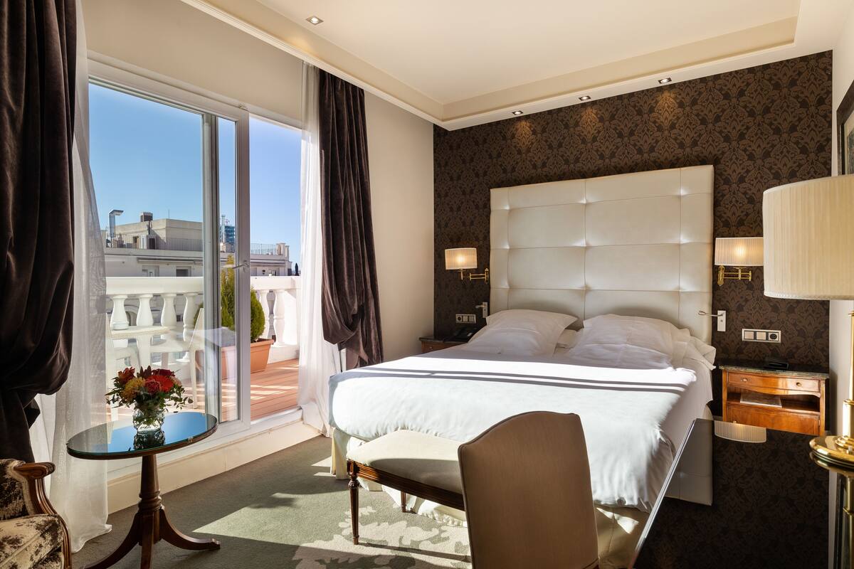 Quarto de hotel com uma cama grande, mesa de cabeceira, luminária, cadeira, pequena mesa com um vaso de flores e vista da cidade através de grandes janelas.