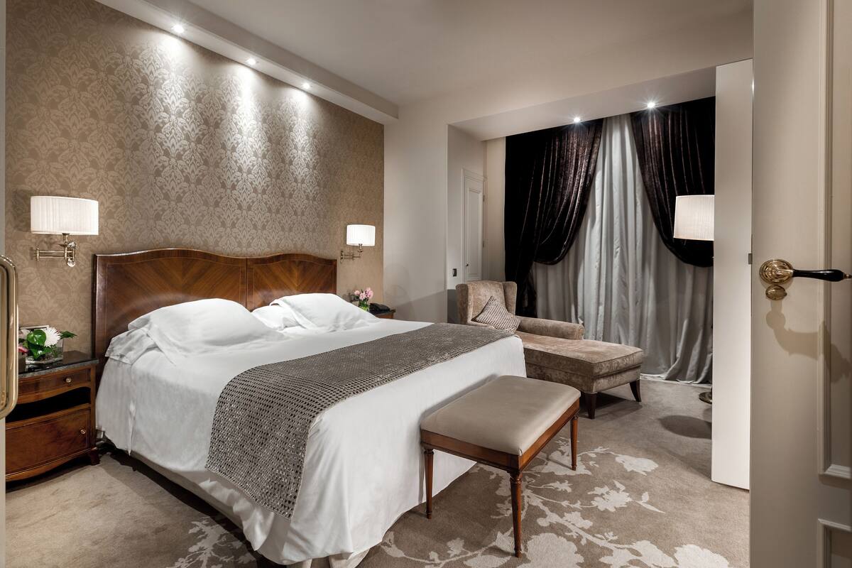 Quarto de hotel com uma cama grande, luminárias de cabeceira, uma cadeira, um banco e piso carpetado.