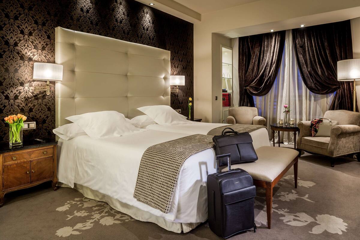 Quarto de hotel com uma cama grande, um espaço de estar com uma cadeira e uma mesa, uma mala e uma mesinha de cabeceira com um vaso de flores.