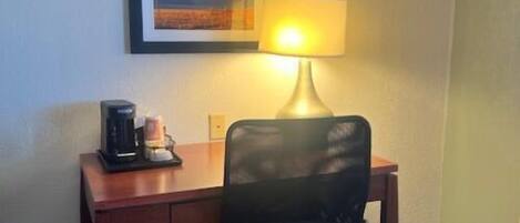 Quarto de hotel com escrivaninha, uma cadeira, luminária, quadro na parede e cafeteira.
