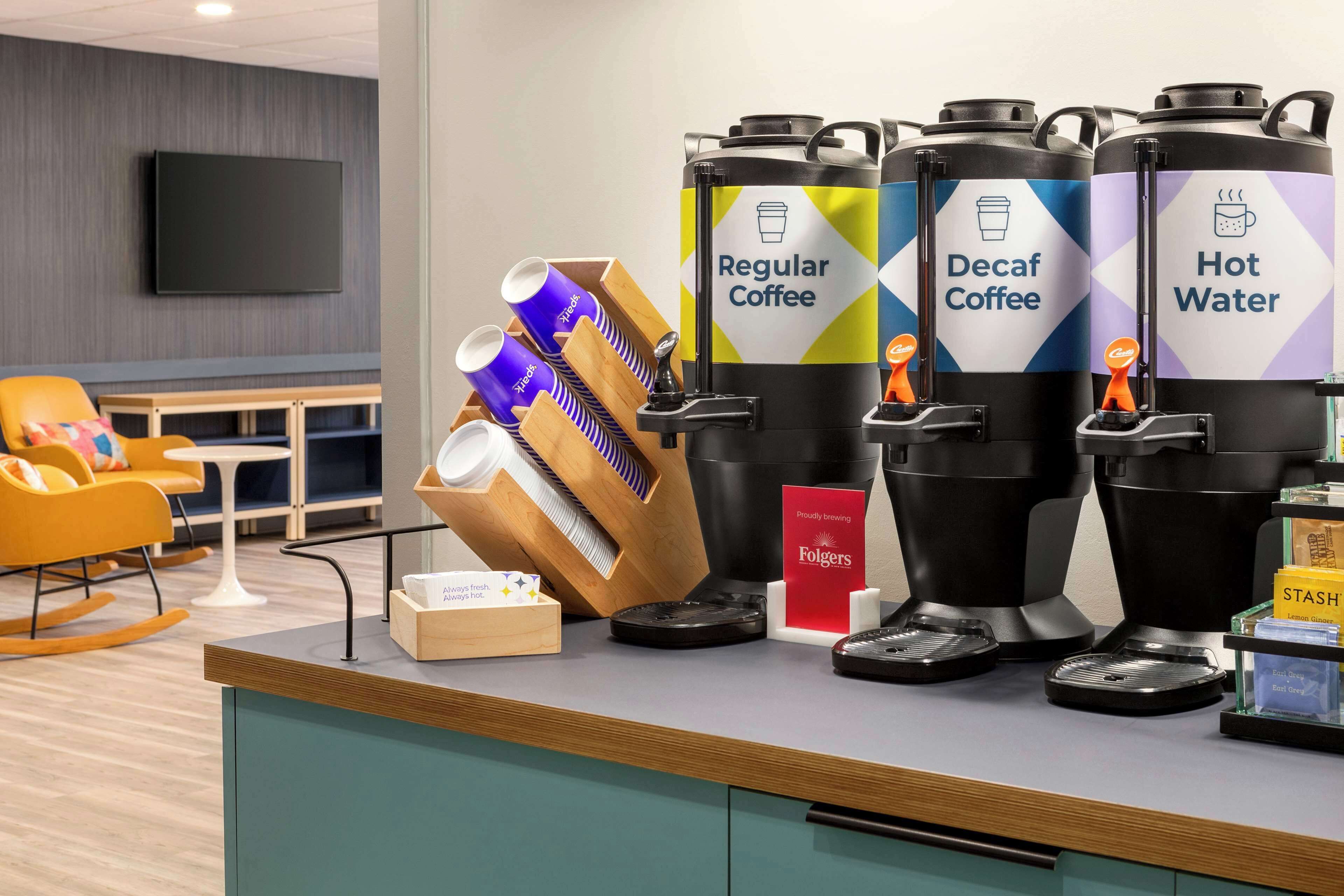 Estação de café com dispensadores para café normal, café descafeinado e água quente, além de uma máquina de café e uma bandeja com copos descartáveis.