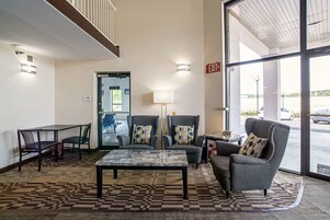Um hall com carpete xadrez, mesa de centro de mármore, duas poltronas com almofadas estampadas, um sofá e uma mesa de jantar com cadeiras.