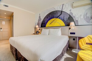 Quarto de hotel com uma cama grande, uma escrivaninha com abajur, uma cadeira e parede com design circular.