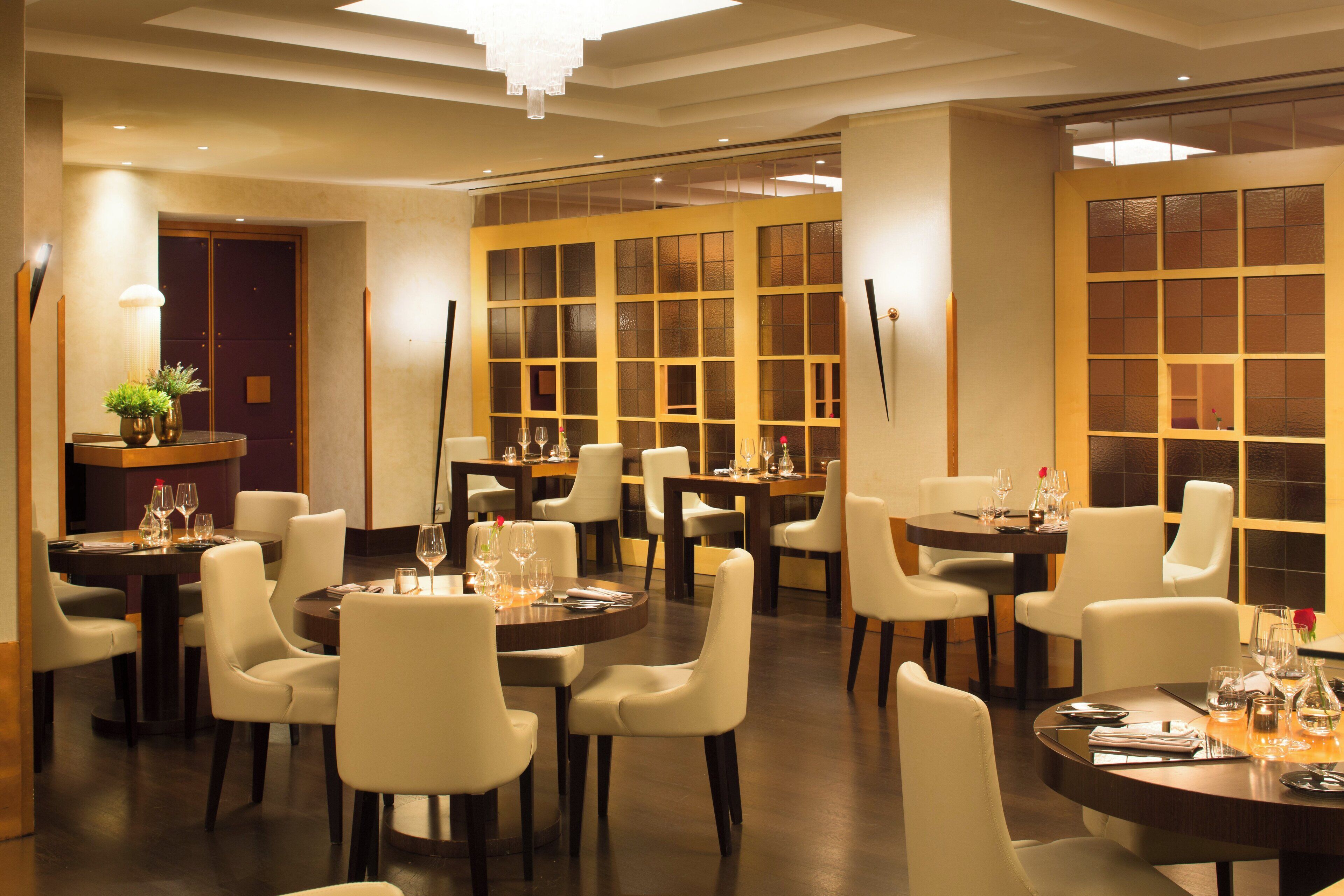 Restaurante com mesas redondas e cadeiras brancas, com interior acolhedor e grandes janelas.