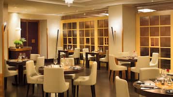 Restaurante com mesas redondas e cadeiras brancas, com interior acolhedor e grandes janelas.