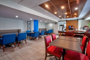 Uma sala de jantar moderna com assentos em azul e vermelho, mesas de madeira e uma cozinha ao fundo.