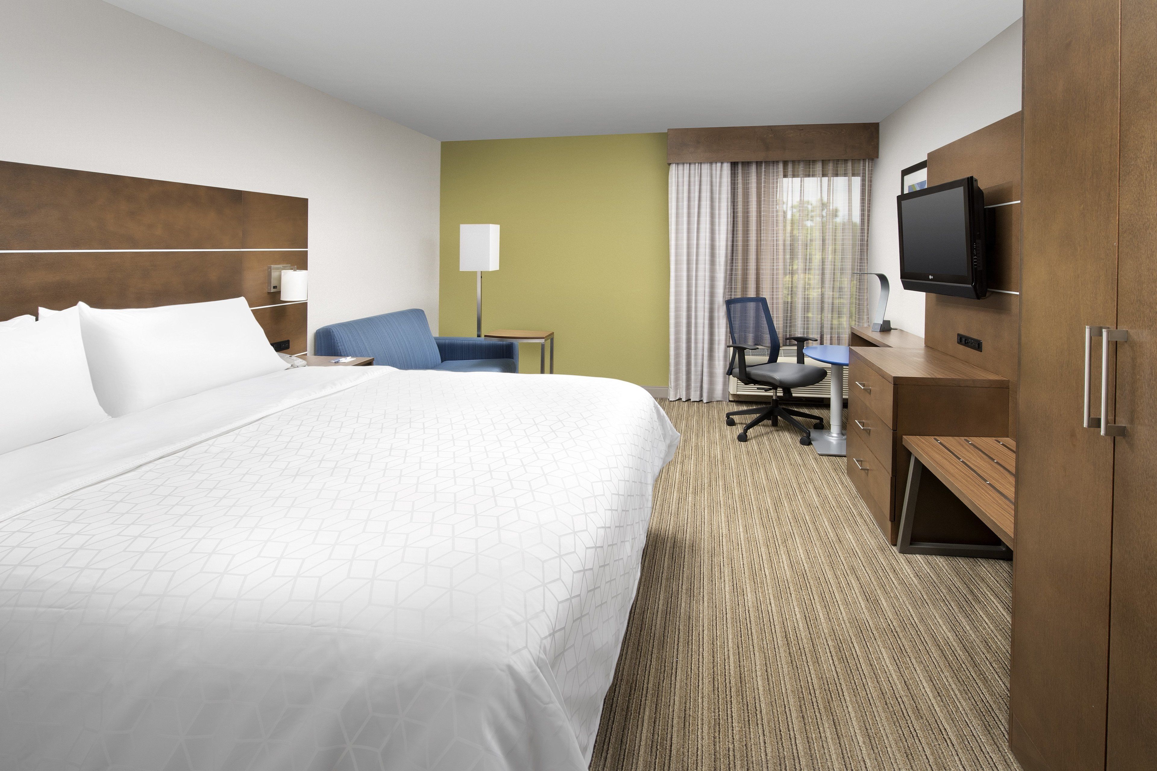 Quarto de hotel com uma cama grande, uma escrivaninha com computador, uma cadeira, uma televisão e um abajur.