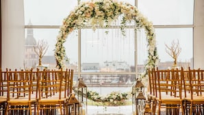 Um espaço interno com um arco de flores e cadeiras de madeira dispostas para um evento.