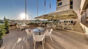 Um pátio externo com cadeiras e mesas brancas, bandeiras e vista para um horizonte urbano.