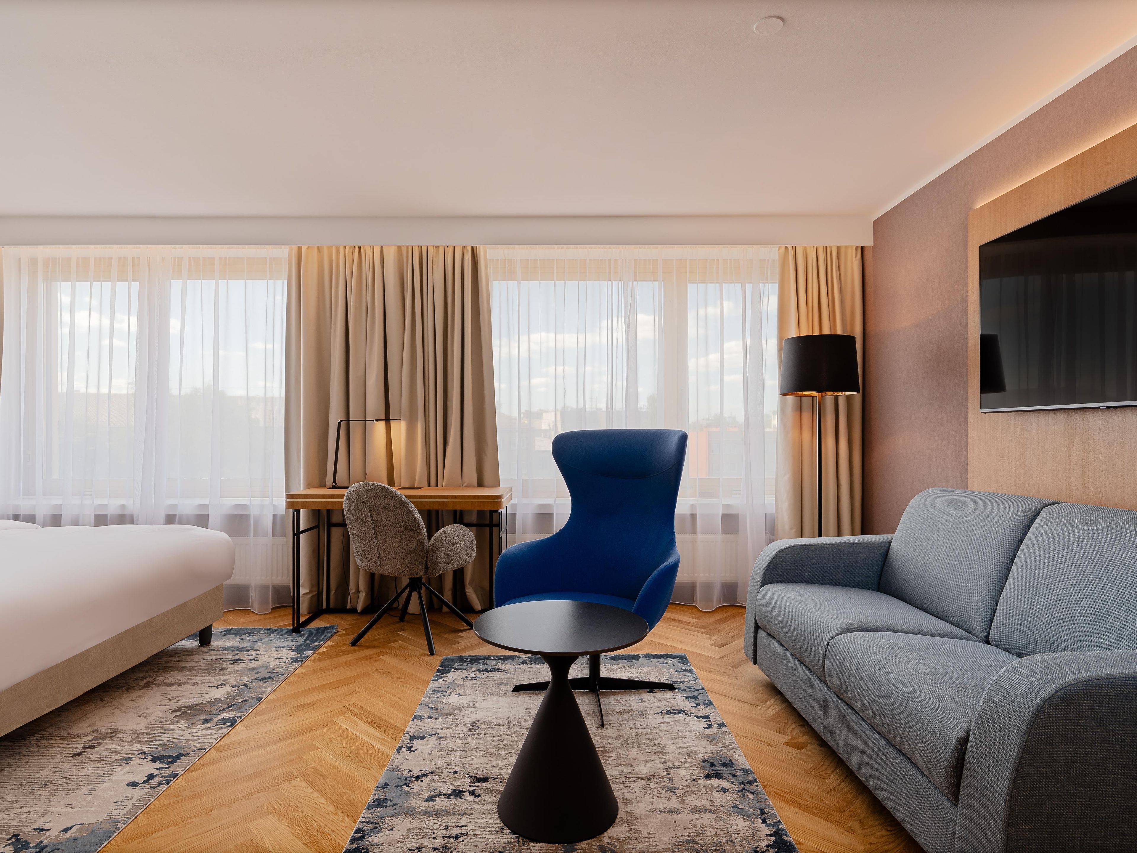 Quarto de hotel com cama, sofá, escrivaninha com cadeira, luminária e televisão.
