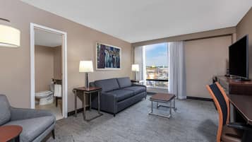 Quarto de hotel com sofá cinzento, uma pequena mesa, uma televisão e vista da cidade através de grandes janelas.