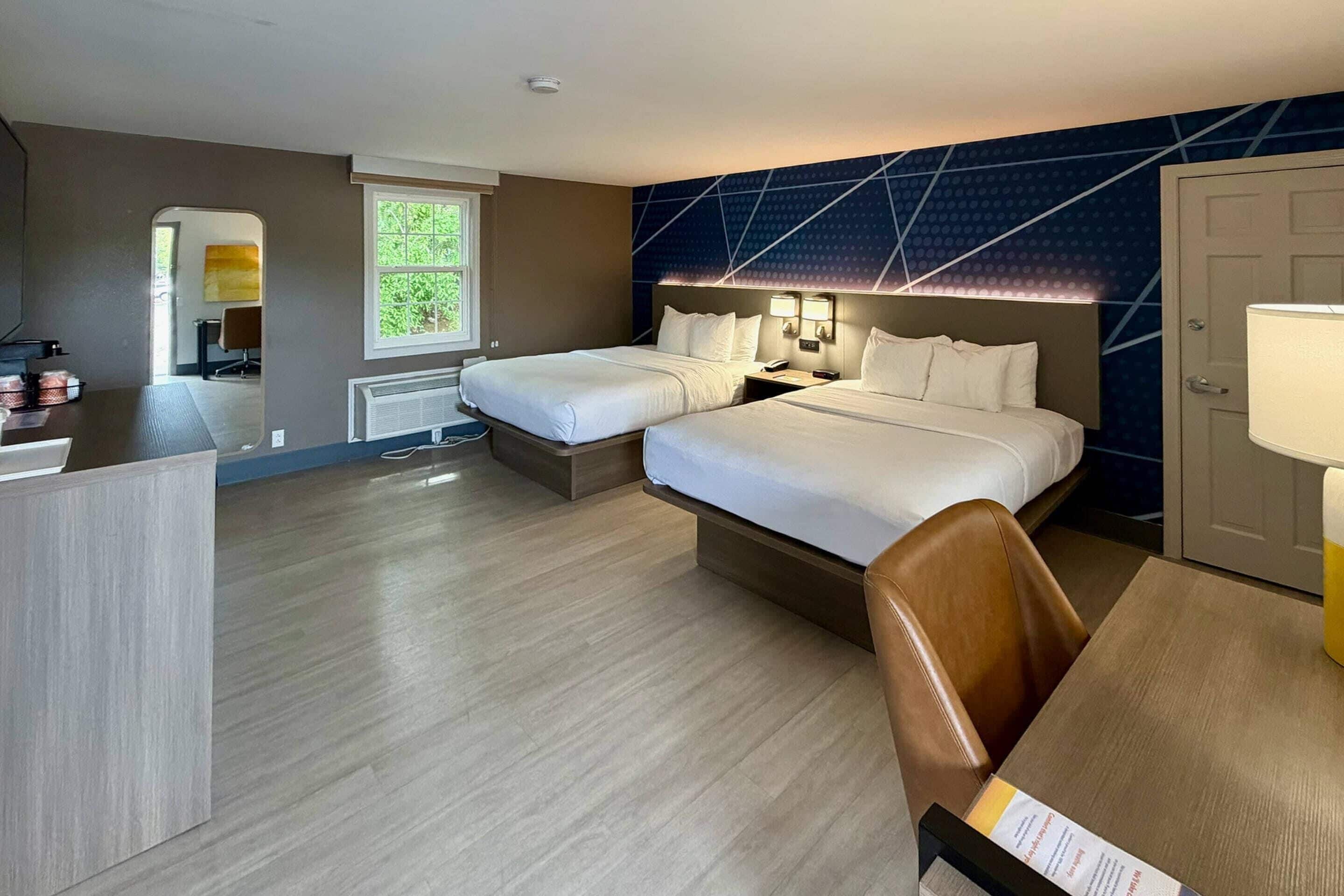 Quarto de hotel com duas camas, uma escrivaninha, uma cadeira, luminária de mesa, mesa de cabeceira, janela e porta.