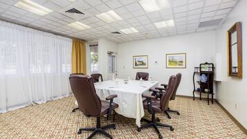 Sala de conferências com uma mesa branca, cadeiras marrons, carpete estampado e uma janela grande com cortinas.