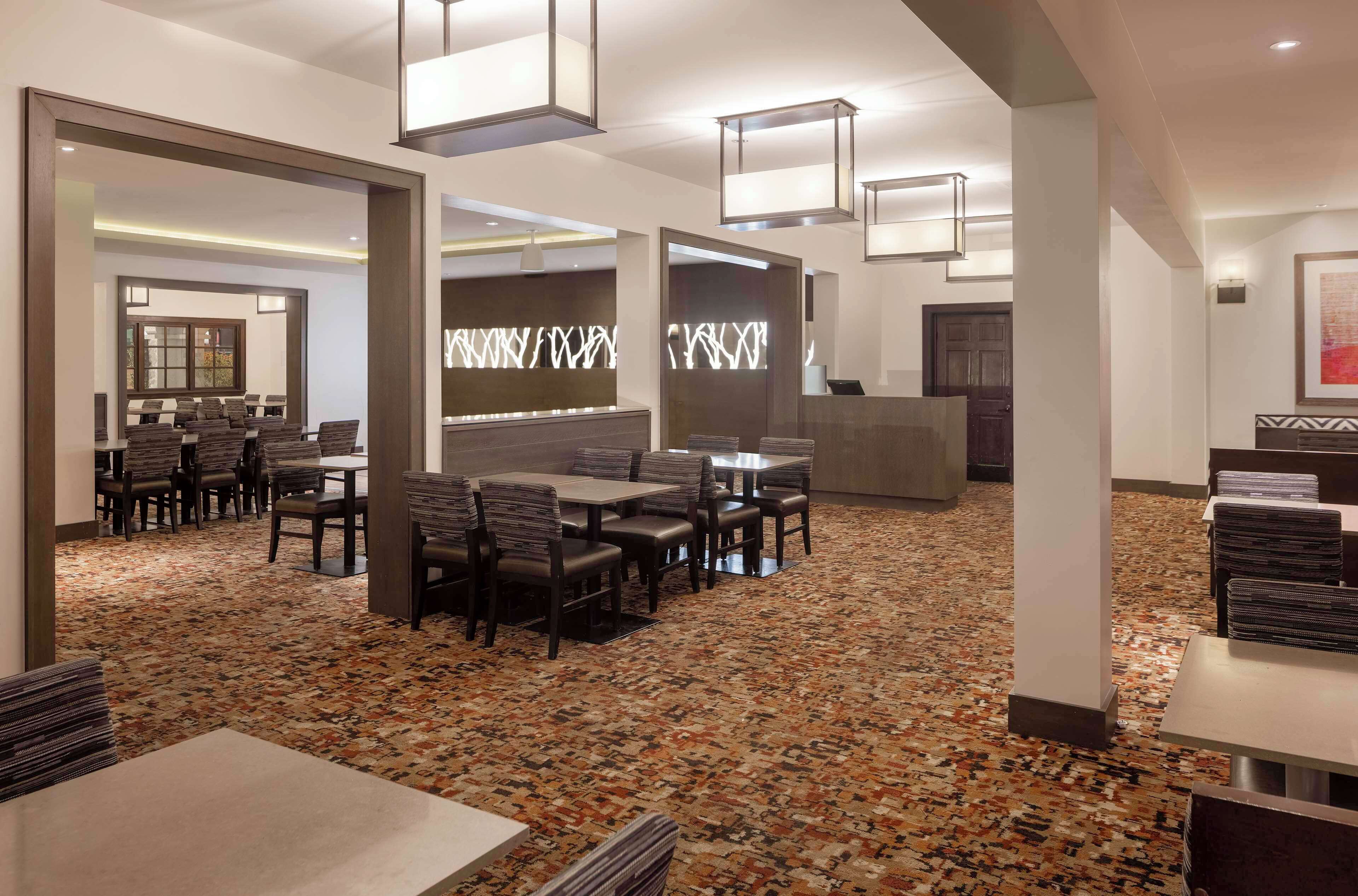 Um saguão de hotel com piso coberto por carpete, várias mesas e cadeiras, e luminárias modernas.