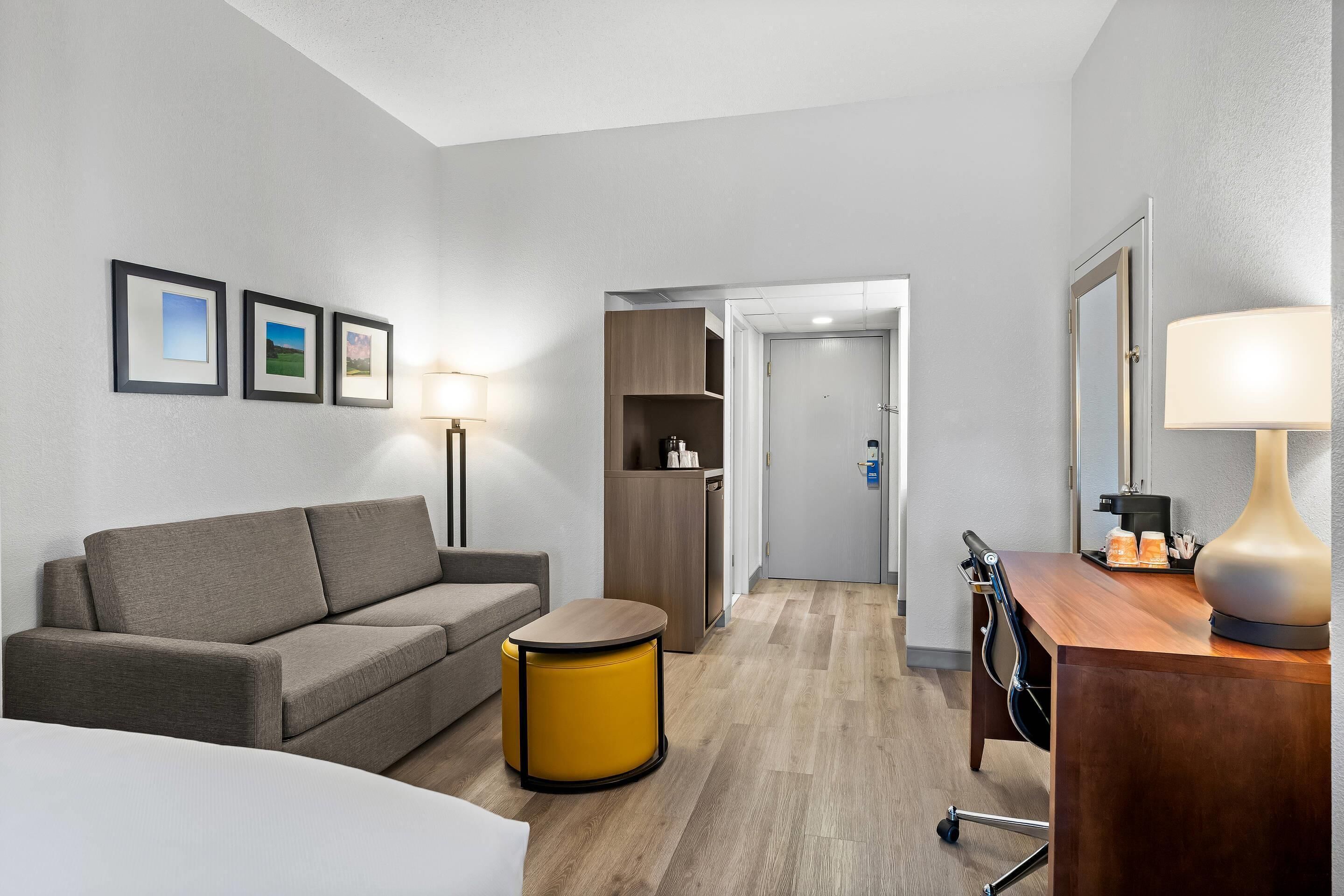 Quarto de hotel com sofá, um banquinho redondo amarelo, secretária de madeira com luminária e quadros nas paredes.