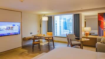 Quarto de hotel com uma cama, uma escrivaninha com cadeira, televisão na parede e uma janela grande com vista para a cidade.