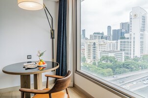 Quarto de hotel com uma mesa redonda, um vaso com flores, uma luminária e vista para a cidade através de uma grande janela.