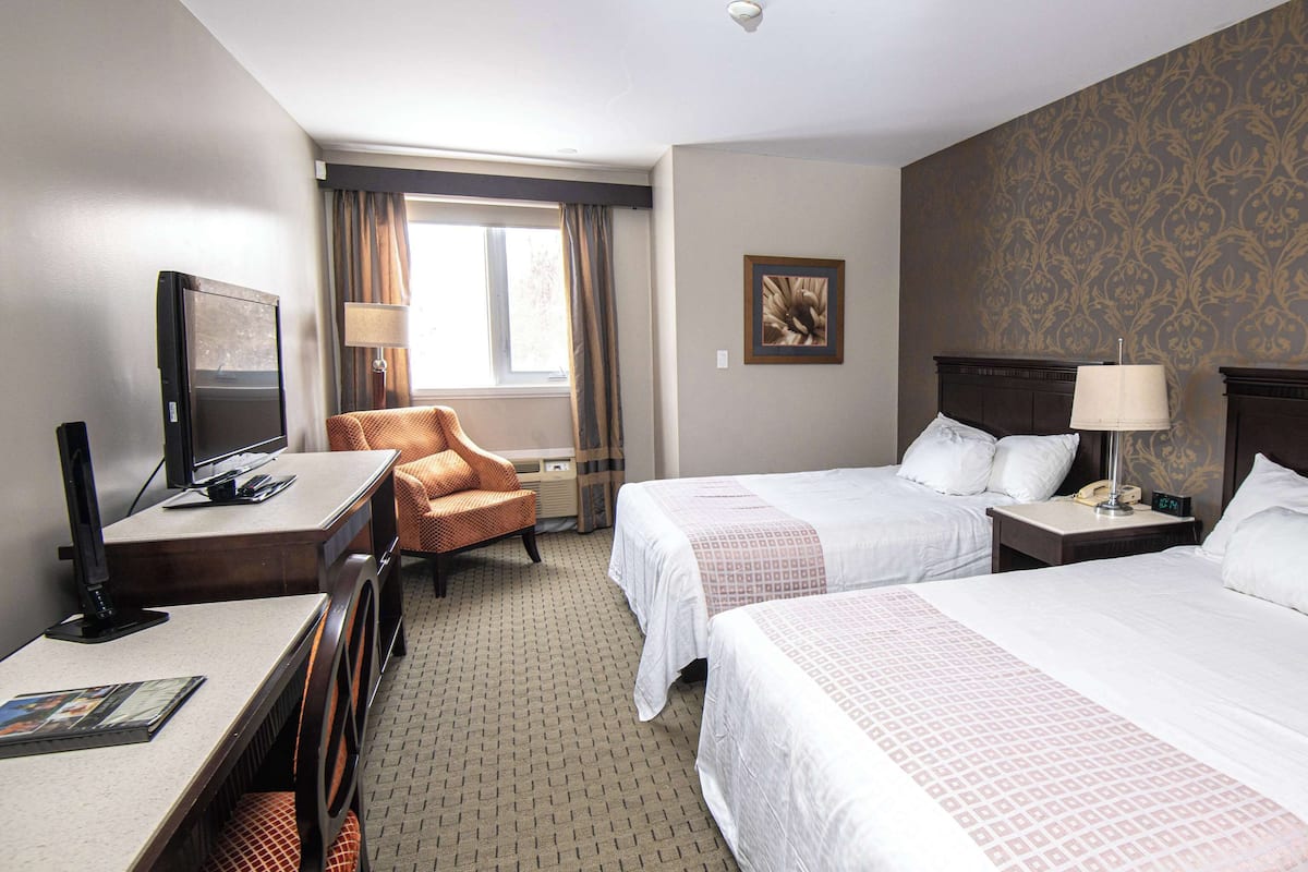 Quarto de hotel com duas camas, uma secretária com computador, uma cadeira, janela com cortinas e parede estampada.