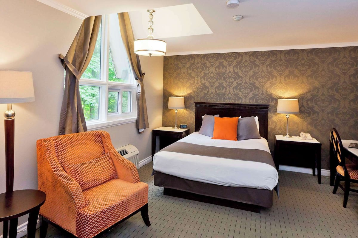 Quarto de hotel com cama, duas mesas de cabeceira, uma cadeira, escrivaninha e janela com cortinas.