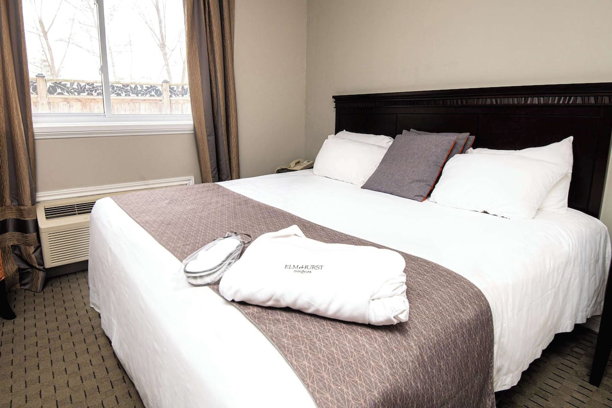 Quarto de hotel com uma cama grande, uma cadeira, mesa de cabeceira e janela com cortinas.