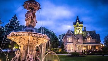 Um edifício histórico com estilo neogótico, uma grande e ornamentada fonte com estátuas e um gramado bem cuidado.