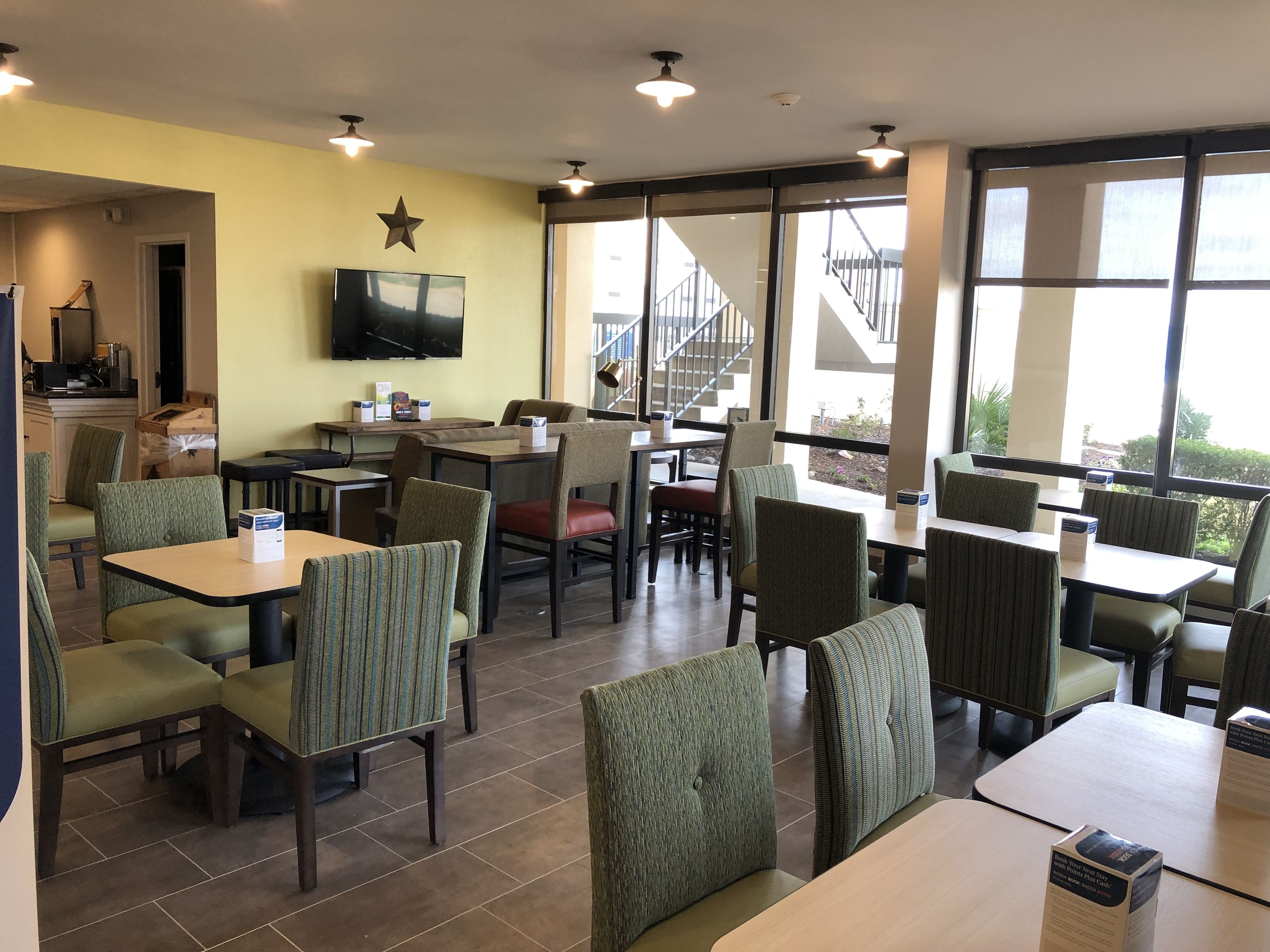 Um restaurante com cadeiras estofadas em verde, mesas, uma televisão de tela plana na parede e janelas amplas com vista para o exterior.