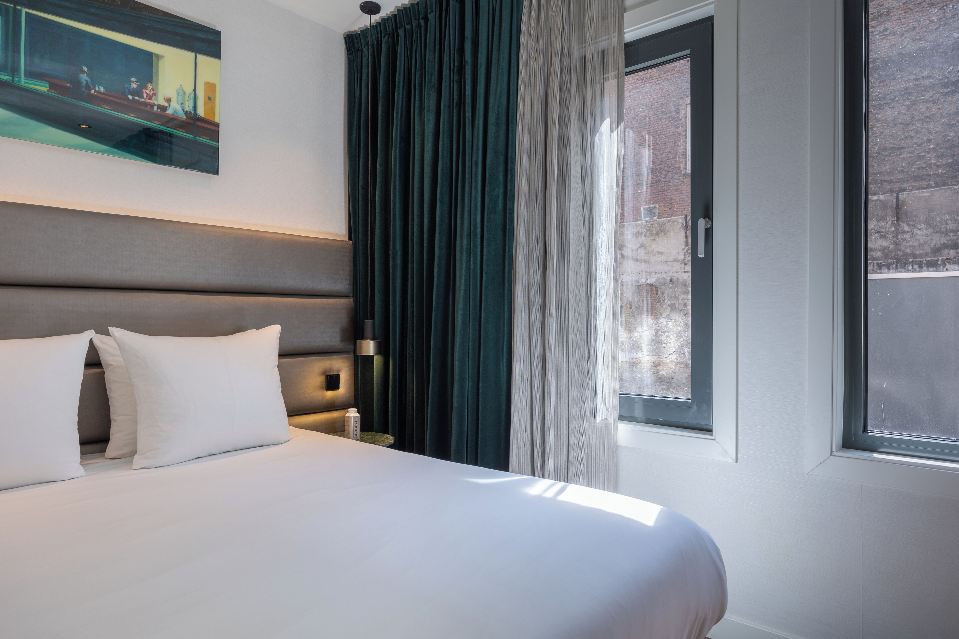 Quarto de hotel moderno com uma cama grande, cabeceira, mesa de cabeceira, pintura na parede e janela com cortinas.