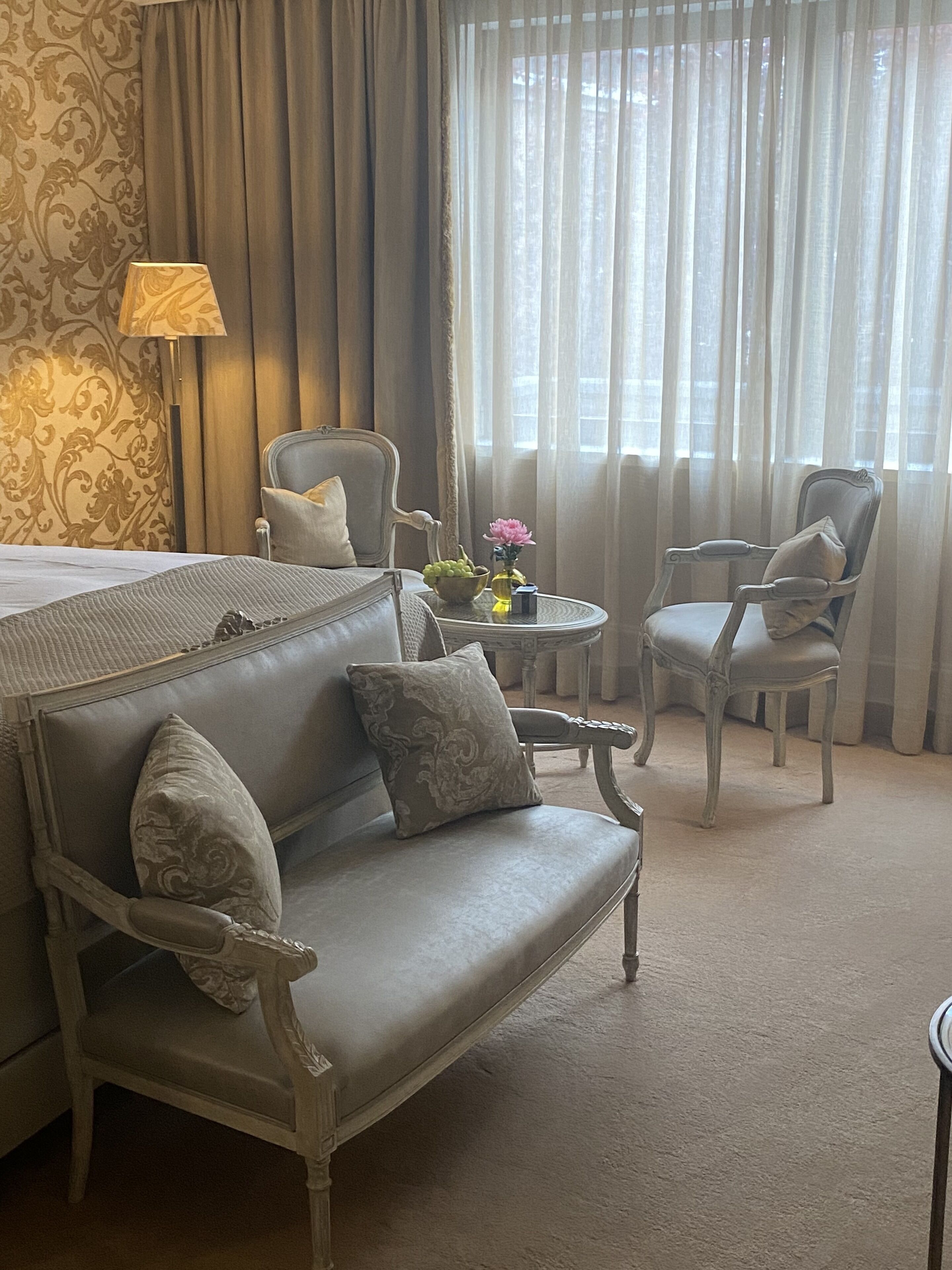 Quarto de hotel com sofá cinzento, duas poltronas, uma mesa pequena com um vaso de flores e um abajur.