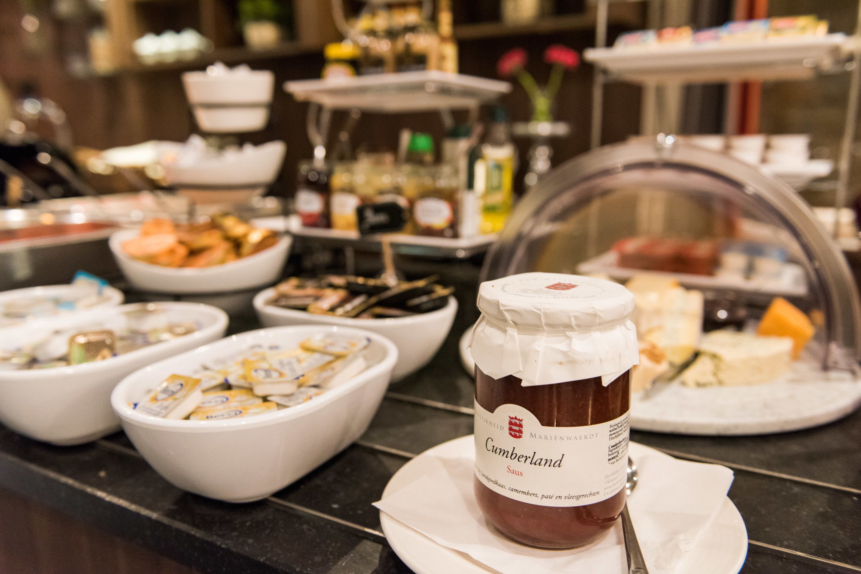 Buffet com diversos tipos de comida, incluindo um pote de Cumberland Saus, recipientes com alimentos e uma vitrine de vidro.
