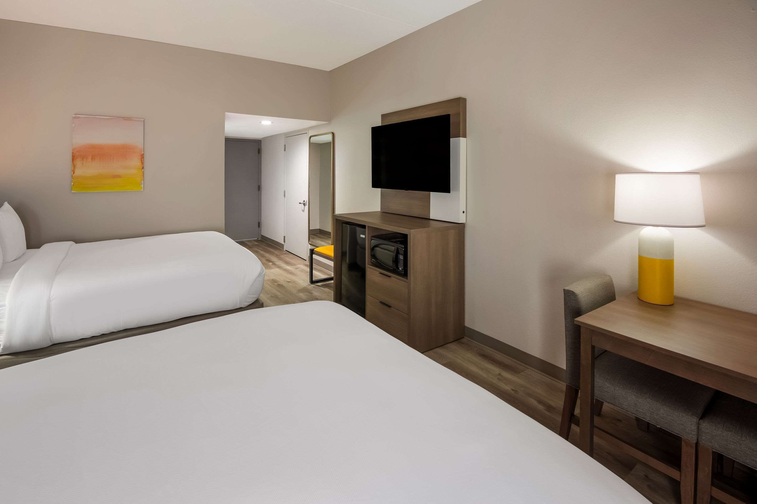 Quarto de hotel com duas camas, televisão, escrivaninha com abajur e um quadro na parede.