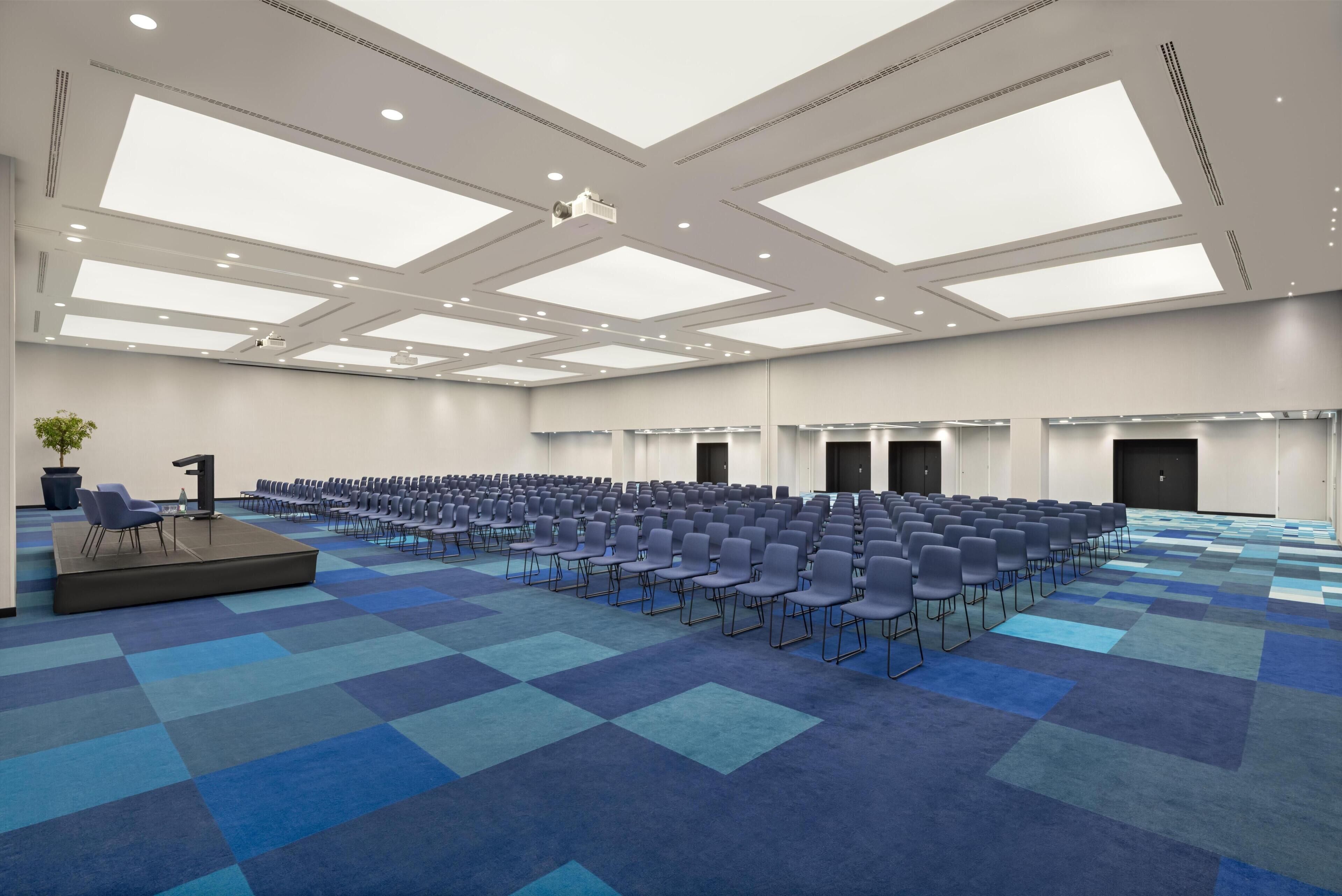 Uma sala de conferências ampla, com fileiras de cadeiras, palco e iluminação embutida no teto.