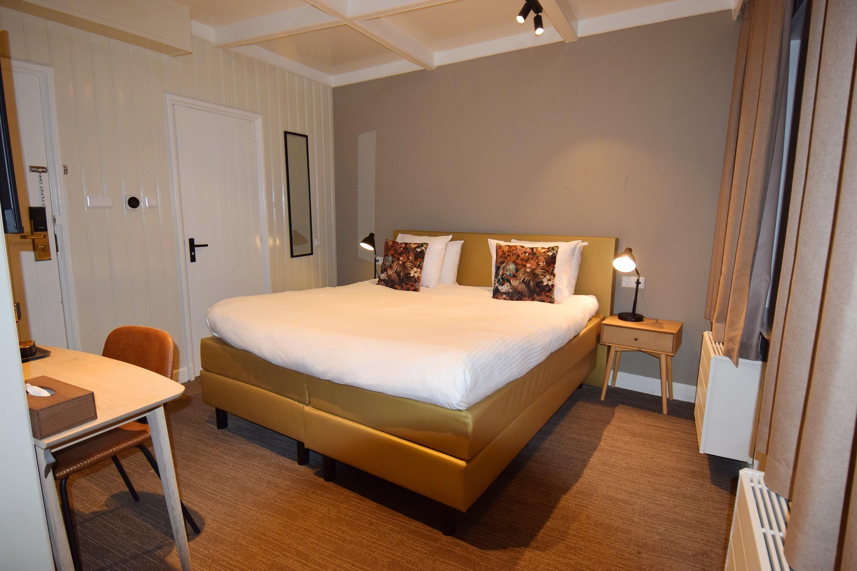 Quarto de hotel com uma cama grande, uma escrivaninha com cadeira, mesa de cabeceira com abajur e radiador.