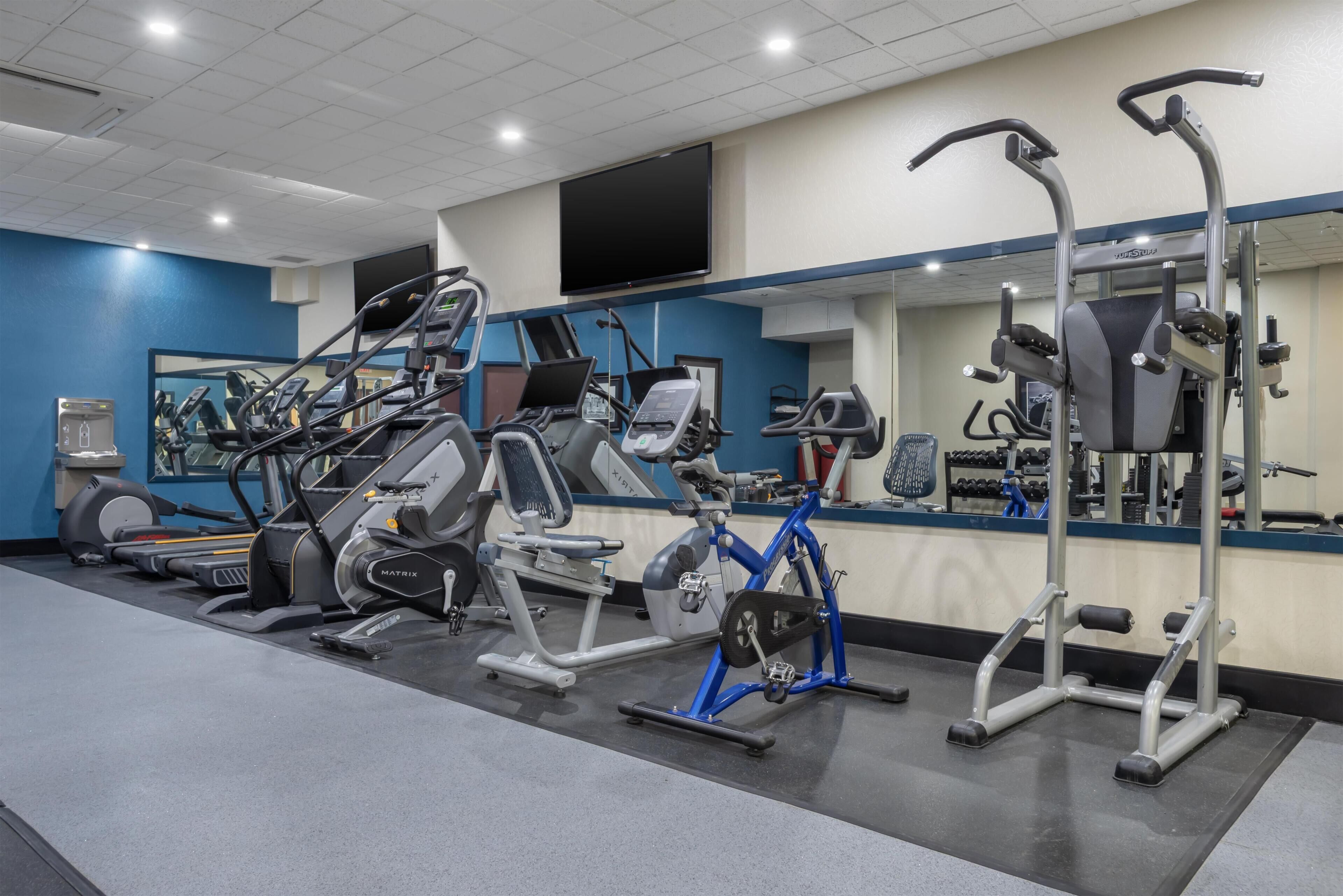Uma academia com diversos equipamentos de exercício, um espelho grande e uma televisão na parede.