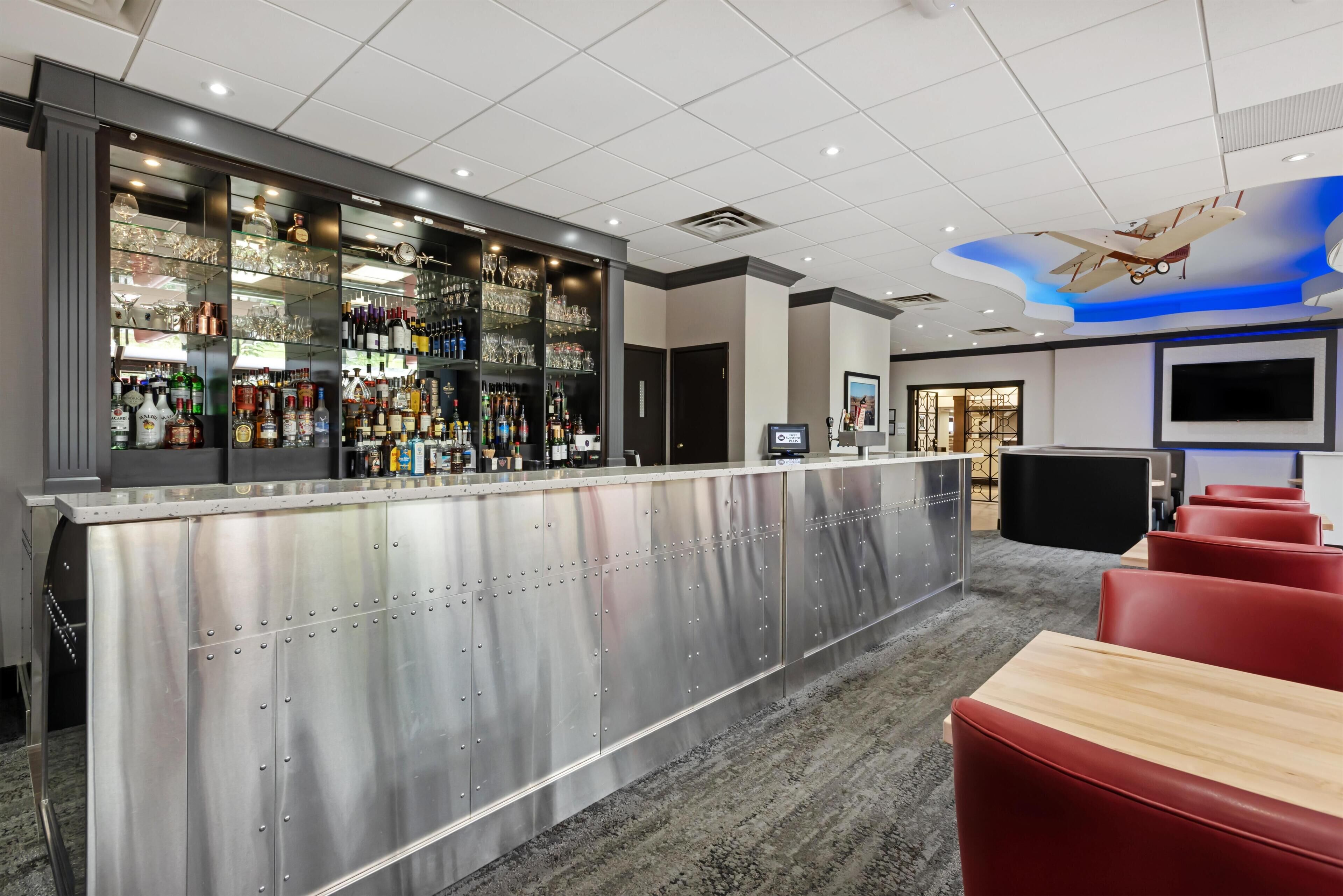 Uma área de bar com balcão de aço inoxidável, prateleiras com garrafas e um espaço de estar com cadeiras vermelhas.