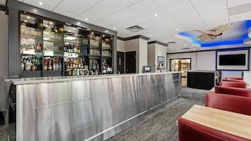 Uma área de bar com balcão de aço inoxidável, prateleiras com garrafas e um espaço de estar com cadeiras vermelhas.