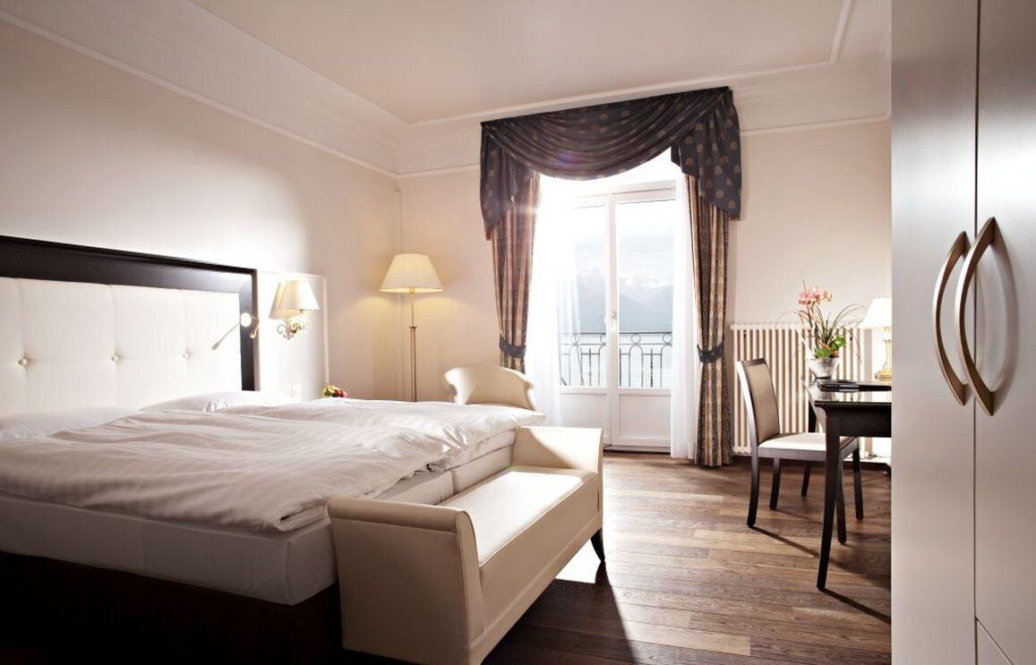 Quarto de hotel com uma cama grande, uma mesa e uma cadeira. Tem uma janela com cortinas e vista para o exterior.
