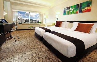 Quarto de hotel com duas camas, uma escrivaninha, uma cadeira, uma mesinha e uma grande janela com vista para a cidade.