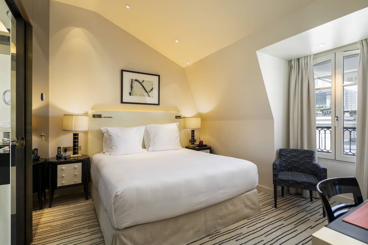 Quarto de hotel com uma cama grande, duas mesas de cabeceira com luminárias, uma cadeira, uma janela com cortinas e um quadro na parede.