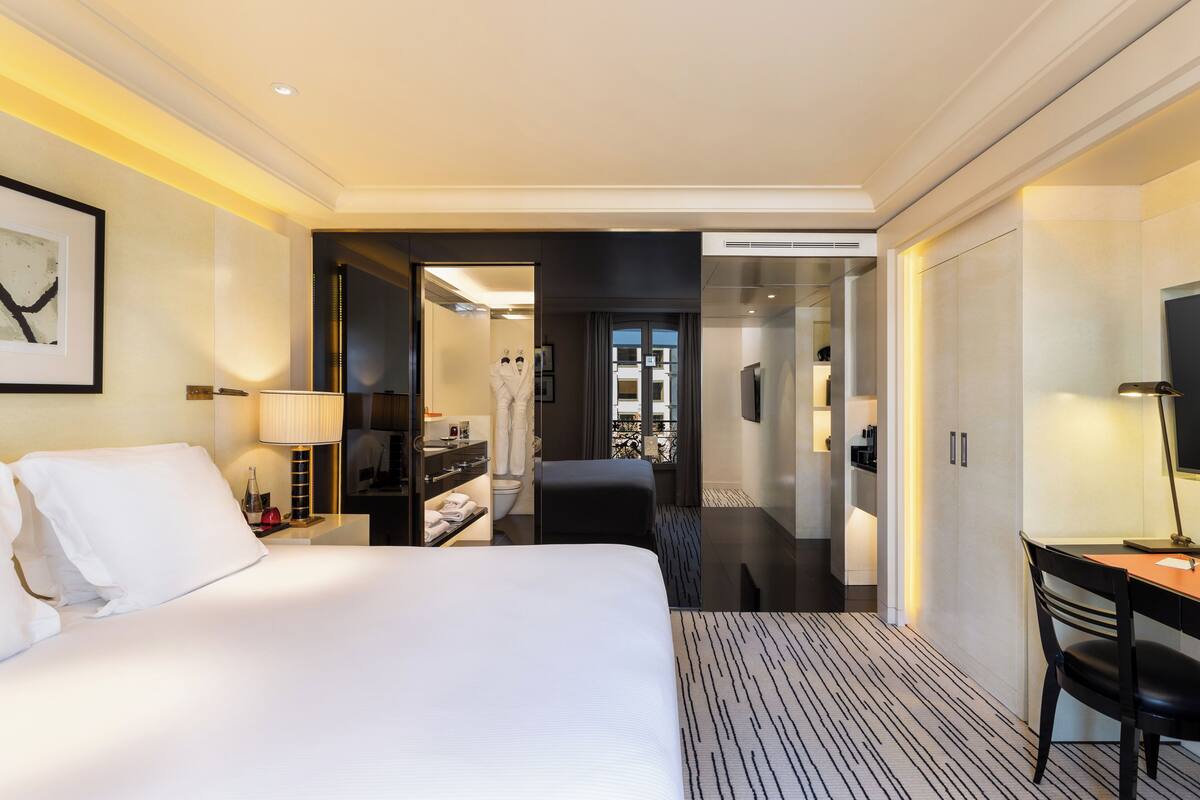 Quarto de hotel com uma cama grande, uma escrivaninha e uma cadeira. Um banheiro é visível por uma porta aberta.