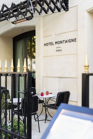 Uma área externa com mesas e cadeiras, uma placa indicando "HOTEL MONTAIGNE PARIS" e uma estrutura metálica decorativa acima.