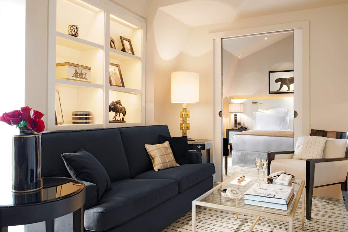 Quarto de hotel moderno com sofá azul escuro, mesa de centro de vidro, estante com fotografias emolduradas, espelho grande e cama com roupa de cama branca.