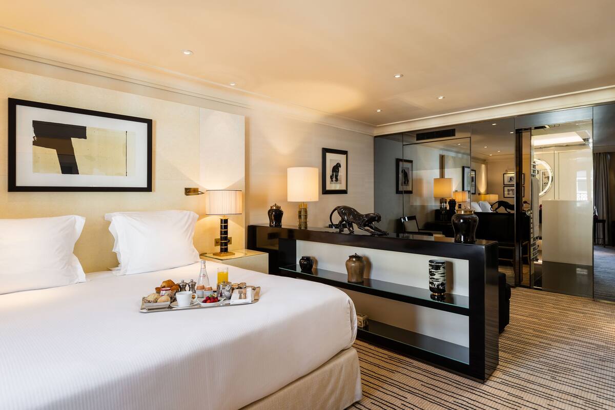 Quarto de hotel com uma cama grande, uma mesa de cabeceira com uma bandeja de itens de café da manhã, uma escrivaninha com abajur e um espelho refletindo o quarto.