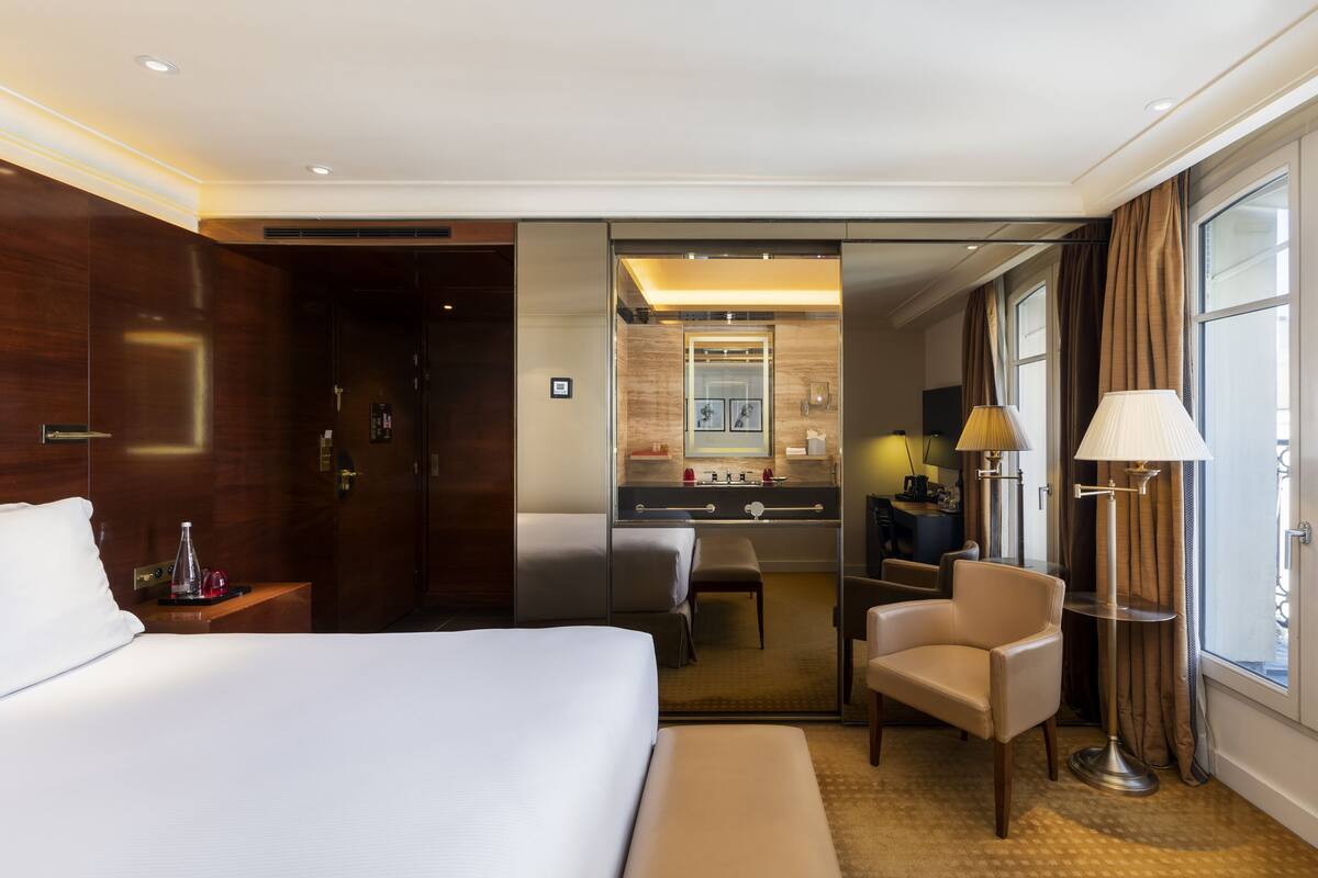 Quarto de hotel com uma cama grande, uma escrivaninha com cadeira, uma mesinha e uma luminária.