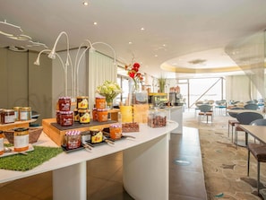 Uma área de recepção de hotel com um balcão exibindo diversos alimentos, uma máquina de café e um espaço de estar com mesas e cadeiras.