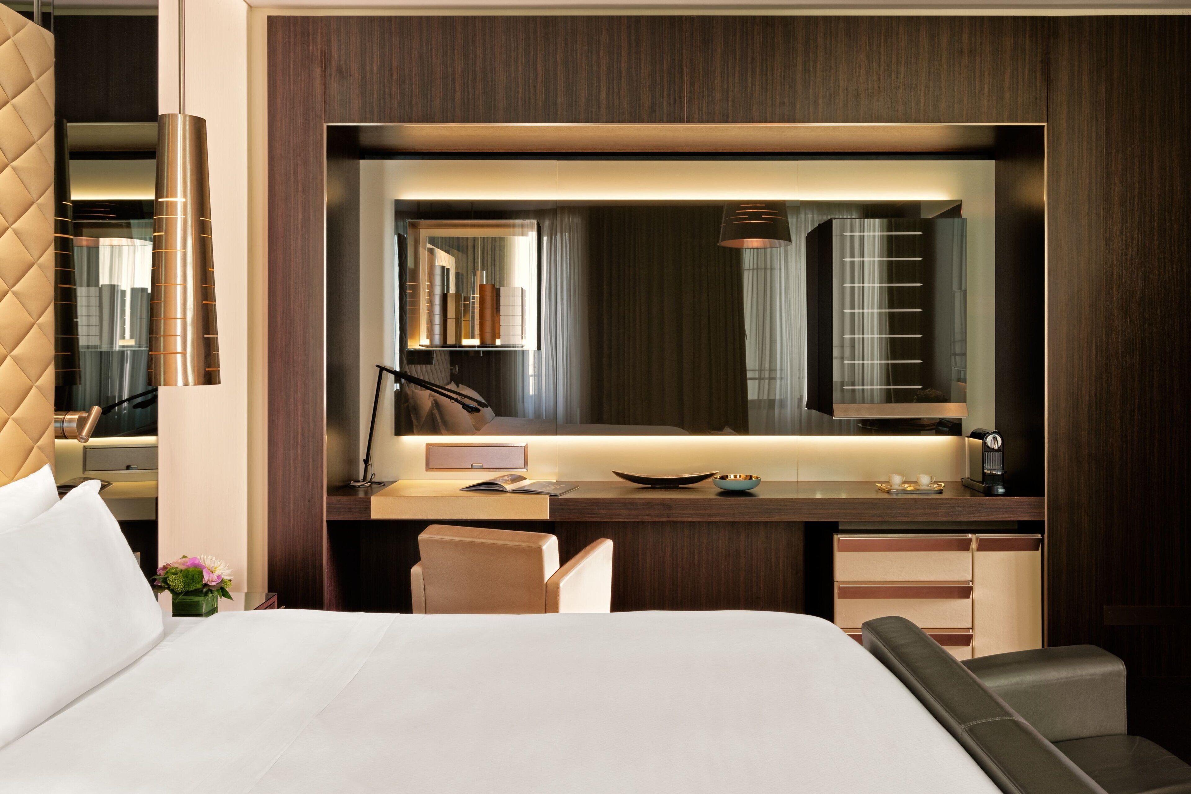 Quarto de hotel moderno com uma cama grande, uma mesa de trabalho com espelho e um espaço para sentar.