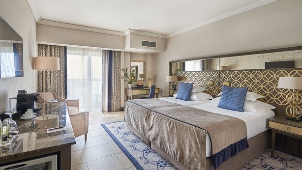 Quarto de hotel com uma cama grande, uma escrivaninha com abajur, uma cadeira e vista para o exterior.
