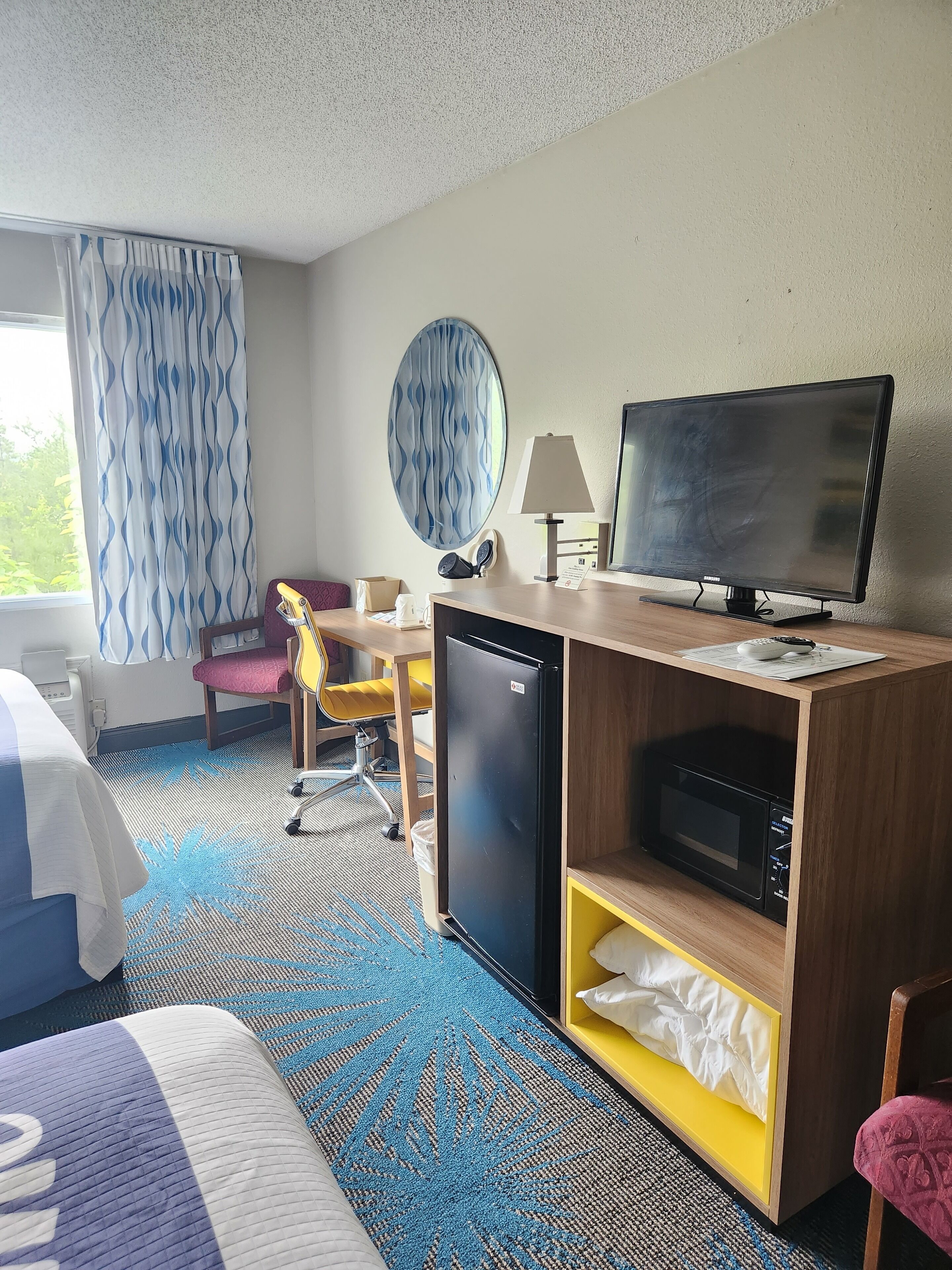 Quarto de hotel com cama, escrivaninha com televisão, micro-ondas, frigorífico, cadeira, abajur e uma janela grande com cortinas.