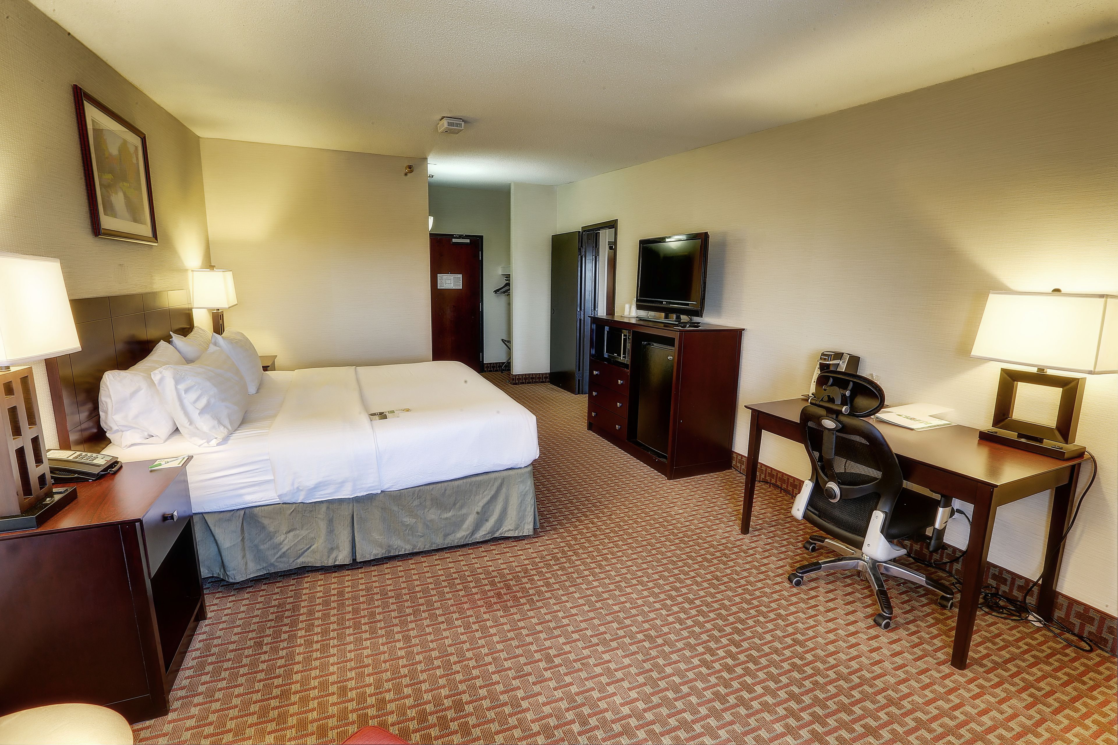 Quarto de hotel com cama, escrivaninha, cadeira, televisão e luminárias.
